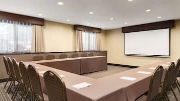 Uma sala de conferências com uma mesa comprida, cadeiras, uma tela de projetor e janelas com cortinas.