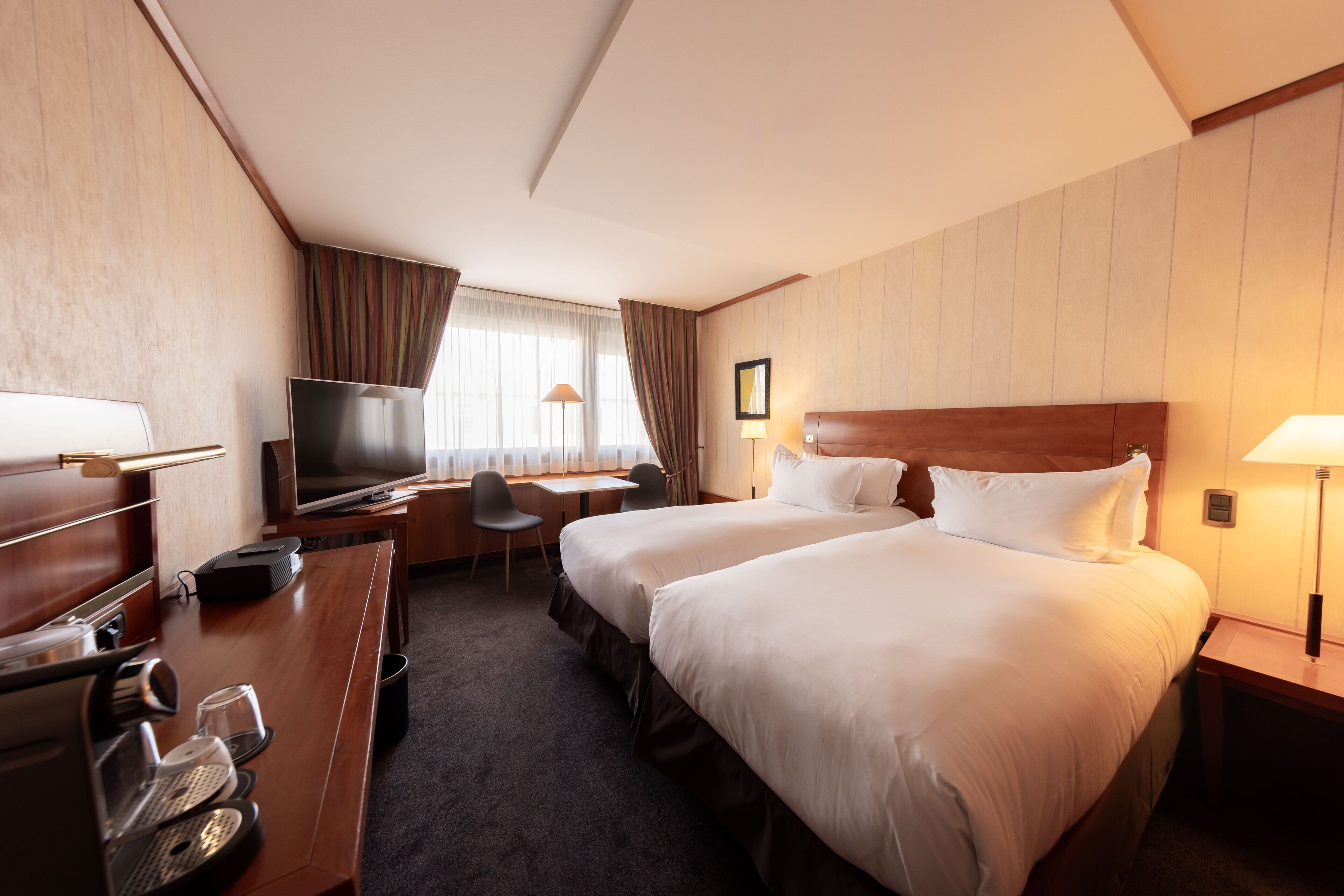 Quarto de hotel com duas camas, uma escrivaninha com televisão e uma cafeteira.