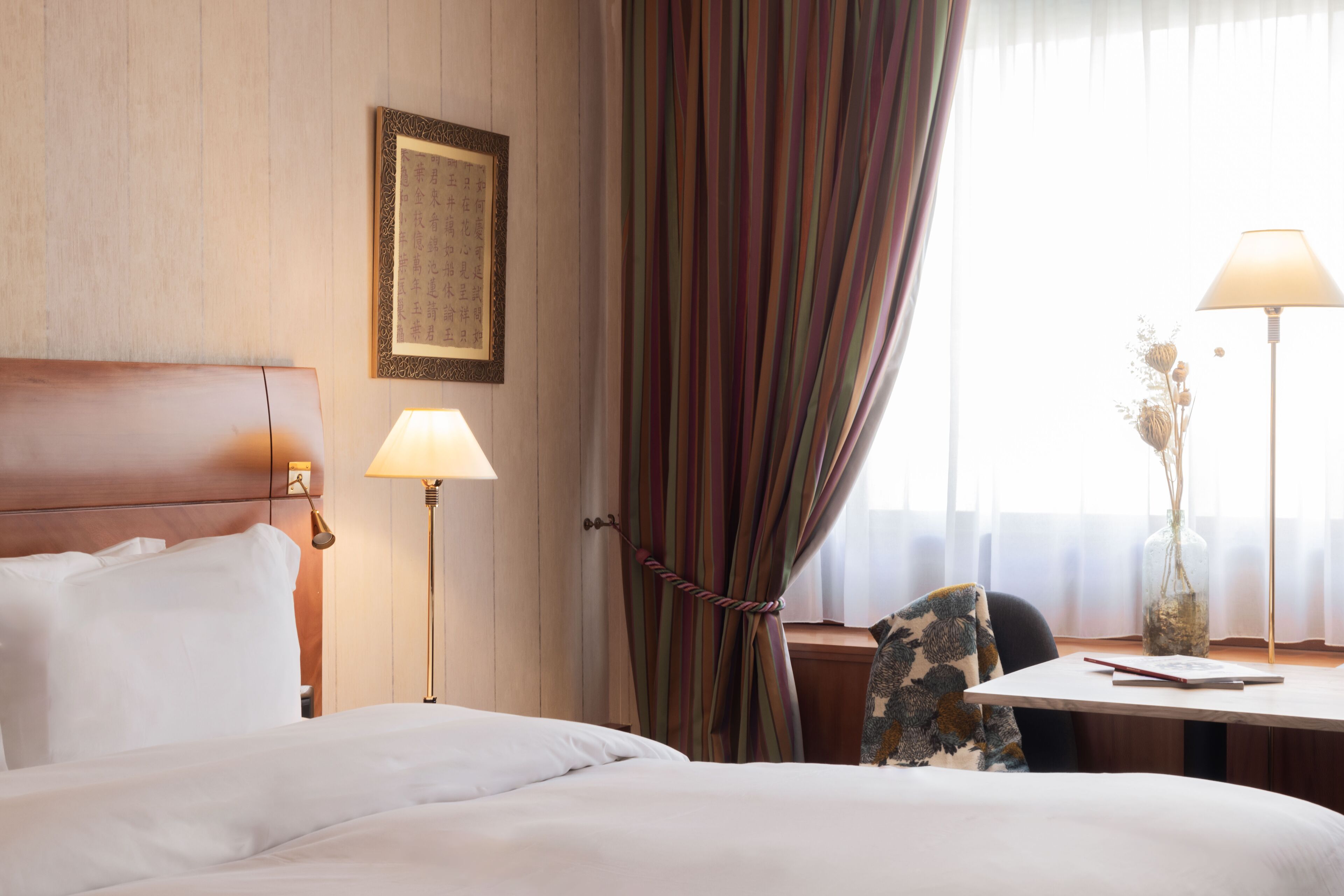 Quarto de hotel com cabeceira de madeira, cama com roupa de cama branca, escrivaninha com luminária, uma cadeira e um vaso com flores secas.