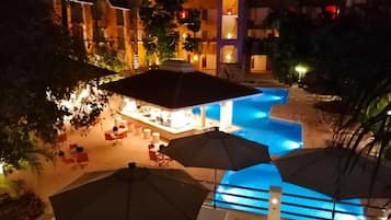Vista noturna de uma área de piscina de hotel, com guarda-sóis, espreguiçadeiras e um bar.