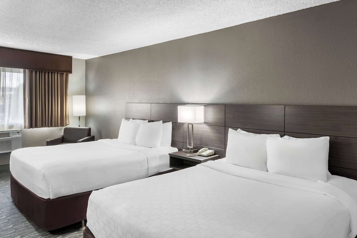 Quarto de hotel com duas camas, uma escrivaninha, uma cadeira e uma luminária.