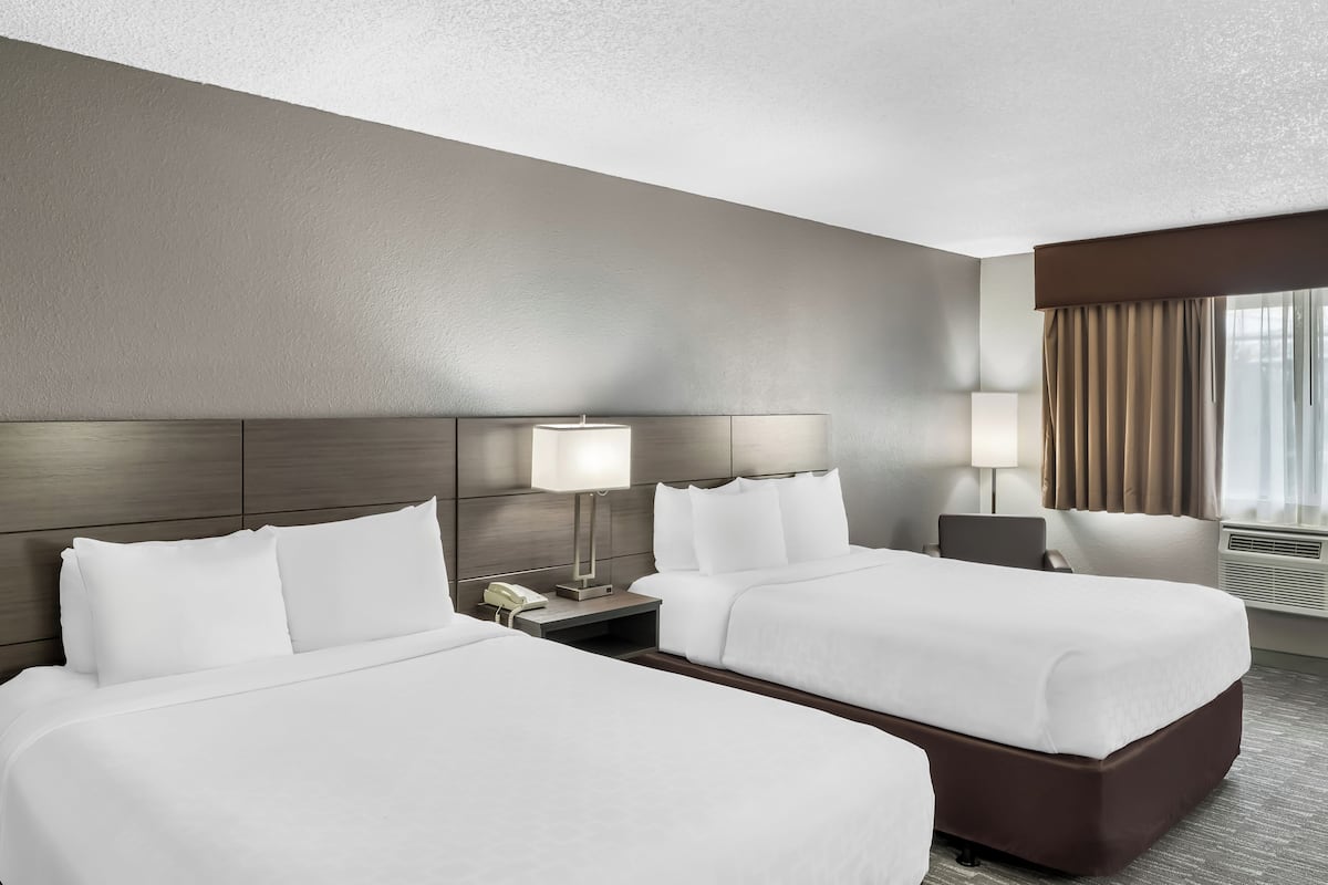 Quarto de hotel com duas camas, uma mesa de cabeceira com abajur, uma cadeira e uma janela com cortinas.