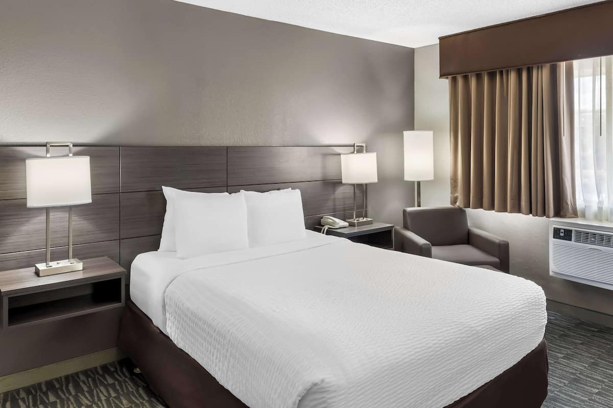 Quarto de hotel com uma cama grande, duas mesas de cabeceira com luminárias, uma cadeira e uma janela com cortinas.