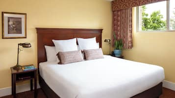 Uma cama bem arrumada com lençóis brancos e almofadas estampadas, uma cabeceira de madeira, uma mesinha de cabeceira com luminária e livros, um quadro na parede e uma janela com cortinas.