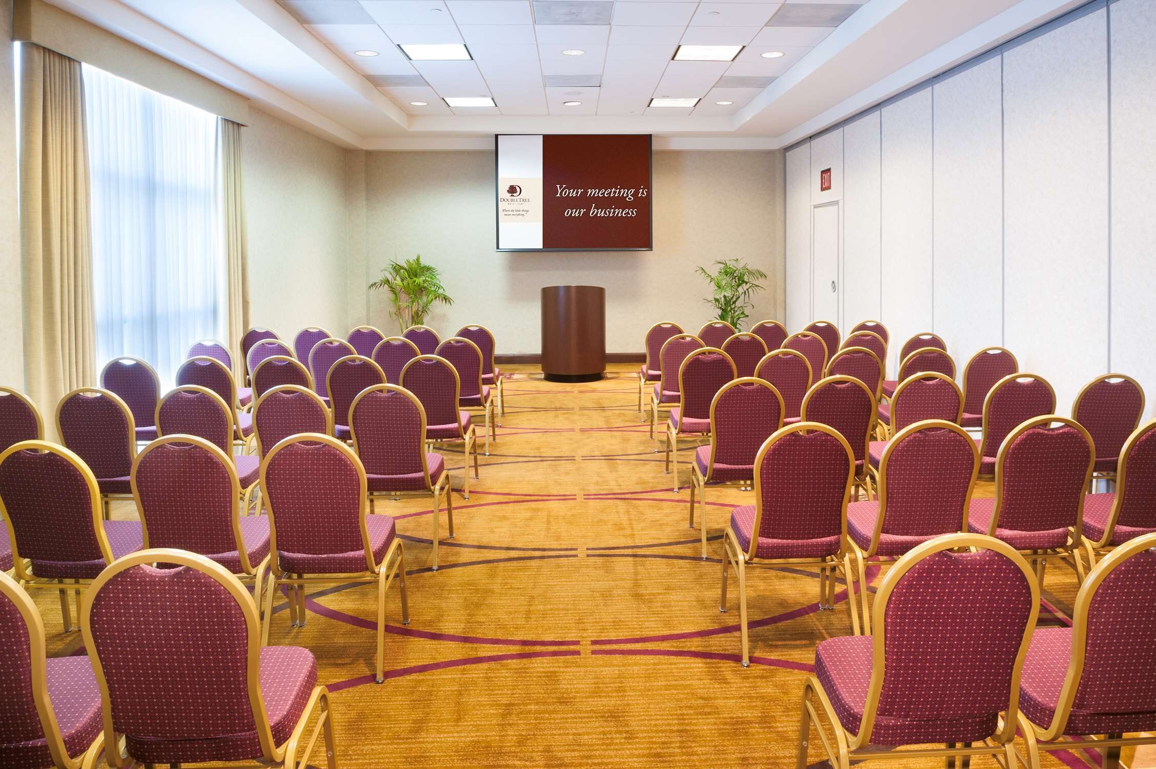 Uma sala de conferências com fileiras de cadeiras roxas, um púlpito e uma tela de projeção.
