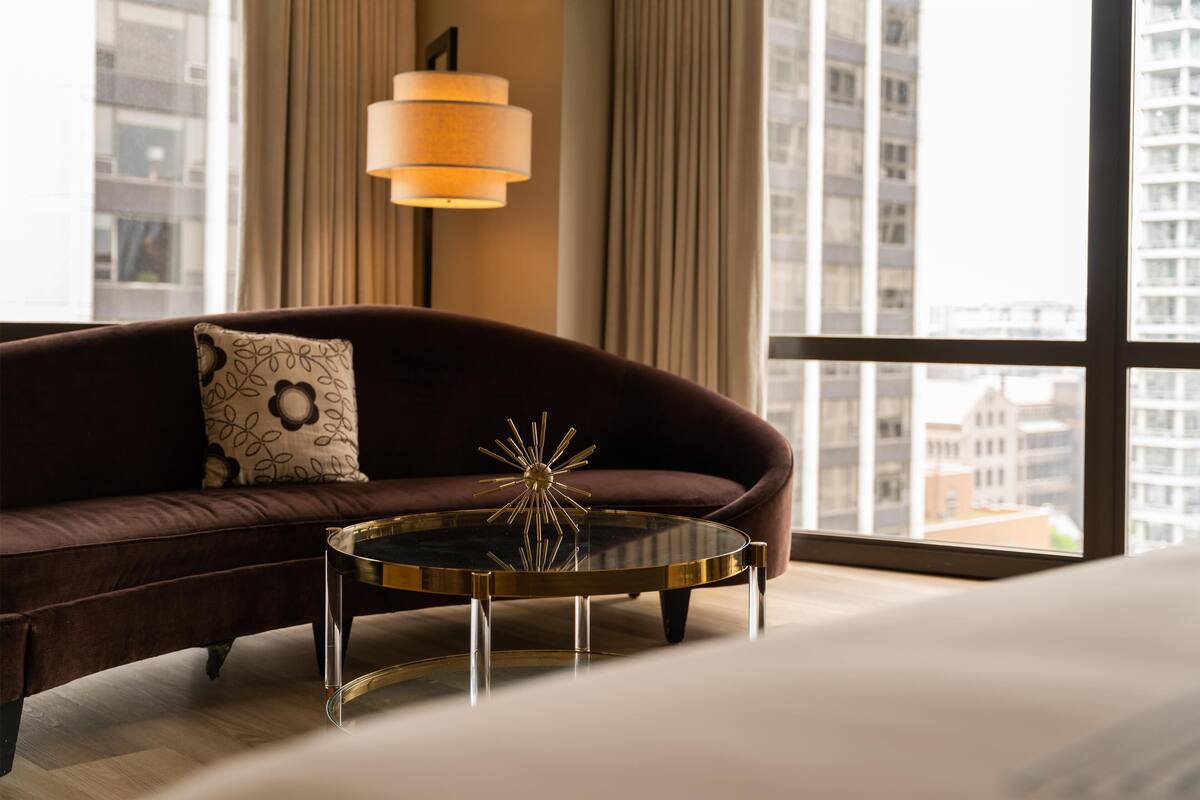 Quarto de hotel moderno com um sofá marrom escuro, uma mesa de centro de vidro com base dourada e uma grande janela com vista para a cidade.