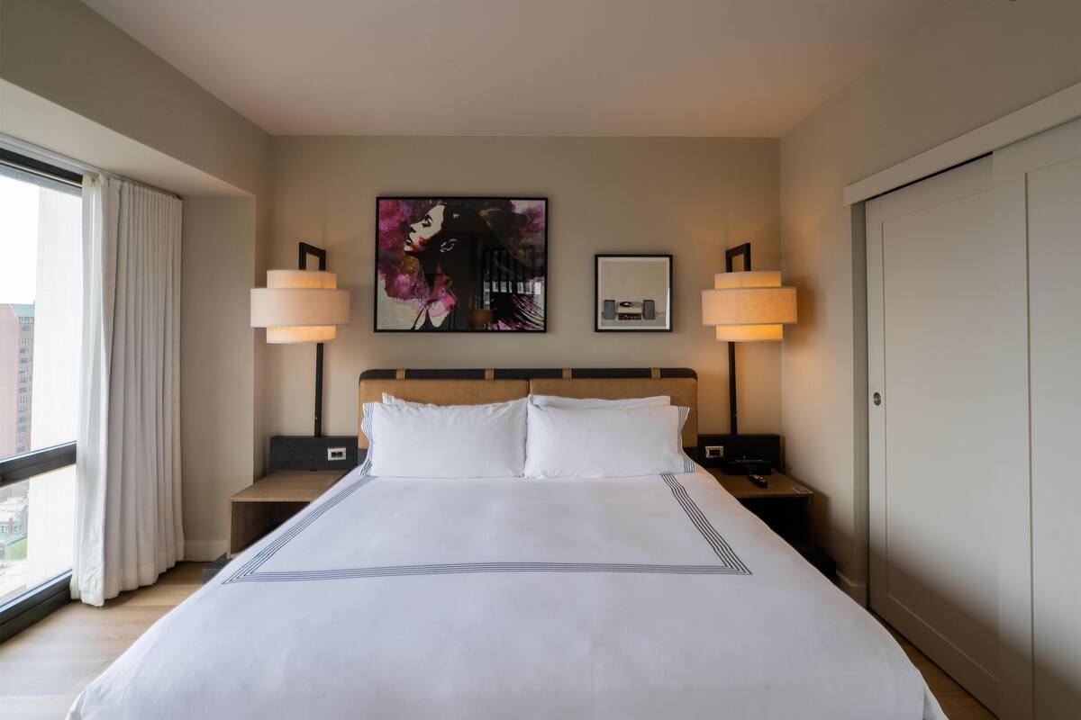Quarto de hotel com uma cama grande, duas luminárias de cabeceira, um quadro na parede e uma janela com cortinas.