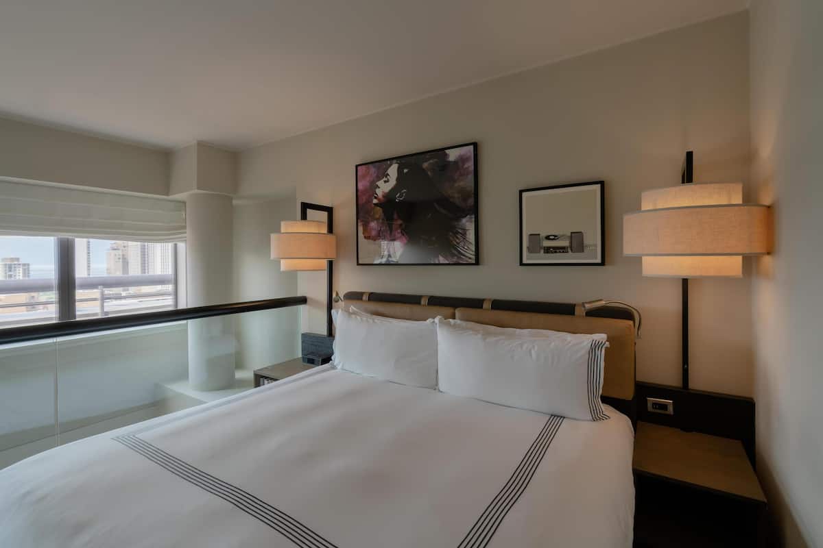 Quarto de hotel com uma cama grande, duas luminárias de cabeceira, um quadro na parede e uma janela com vista para prédios.