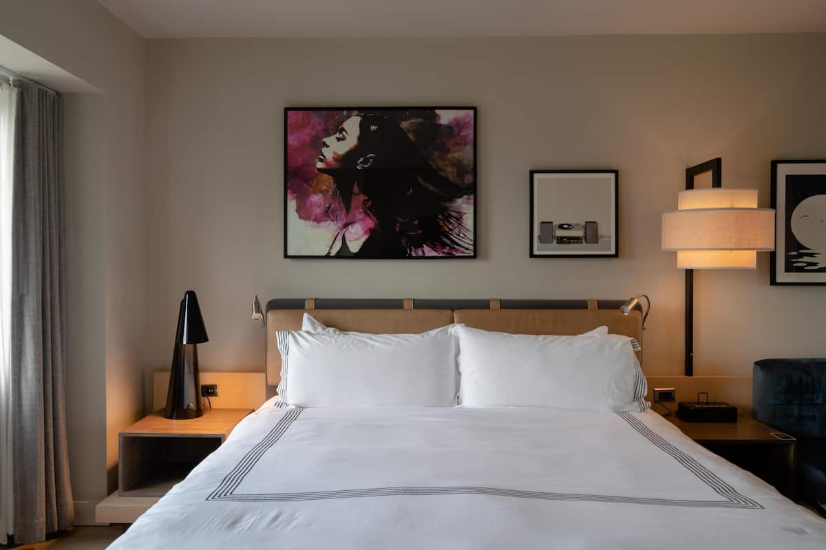 Quarto de hotel com uma cama grande, uma mesa de cabeceira com abajur, um quadro na parede e uma cadeira azul.