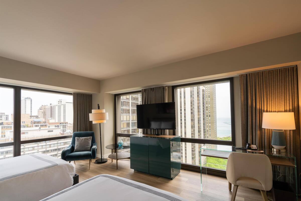 Quarto de hotel moderno com uma cama grande, escrivaninha, cadeira, televisão e vista para a paisagem urbana.