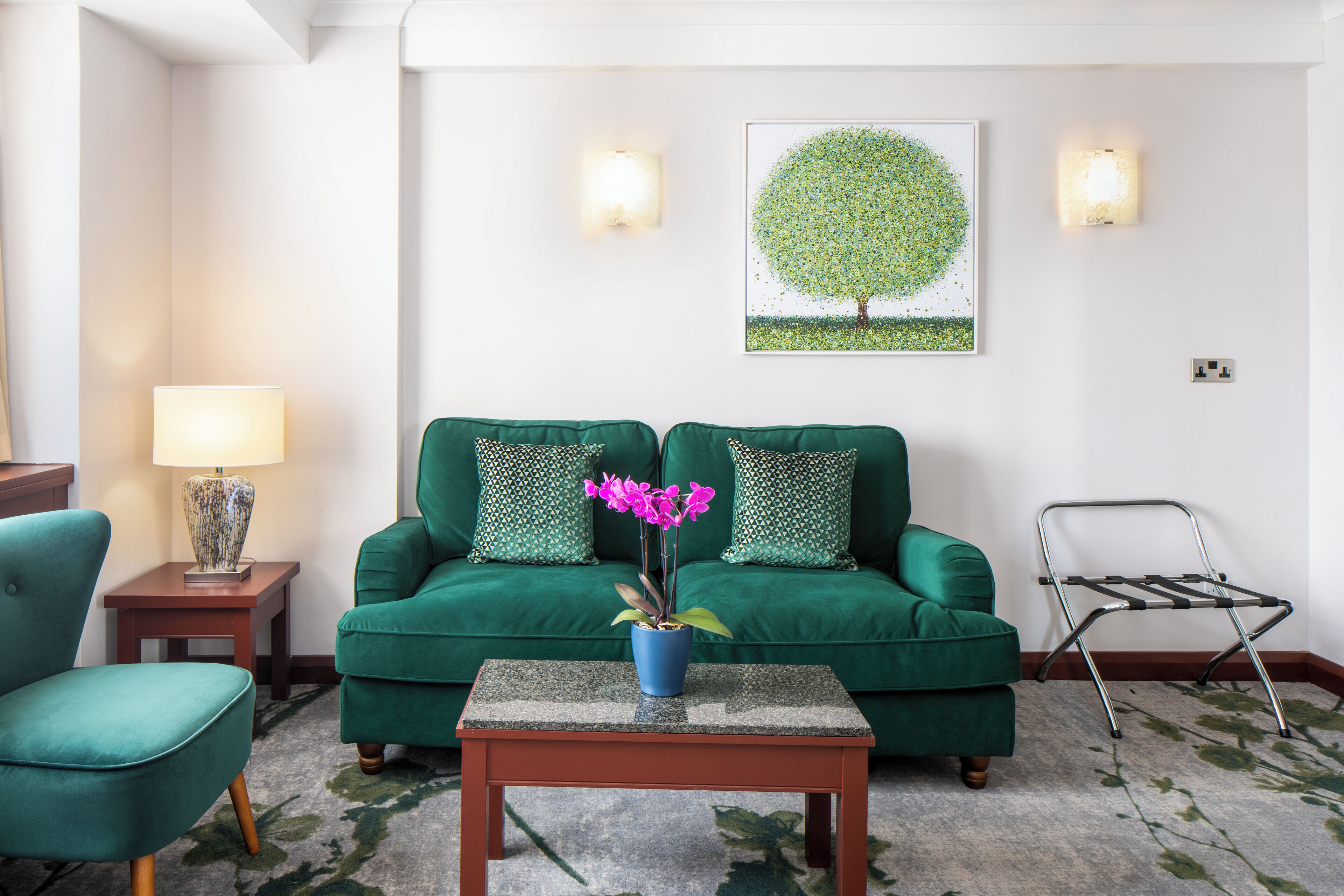 Um sofá verde com almofadas estampadas, uma pequena mesa com um vaso de planta e um quadro de árvore pendurado na parede.