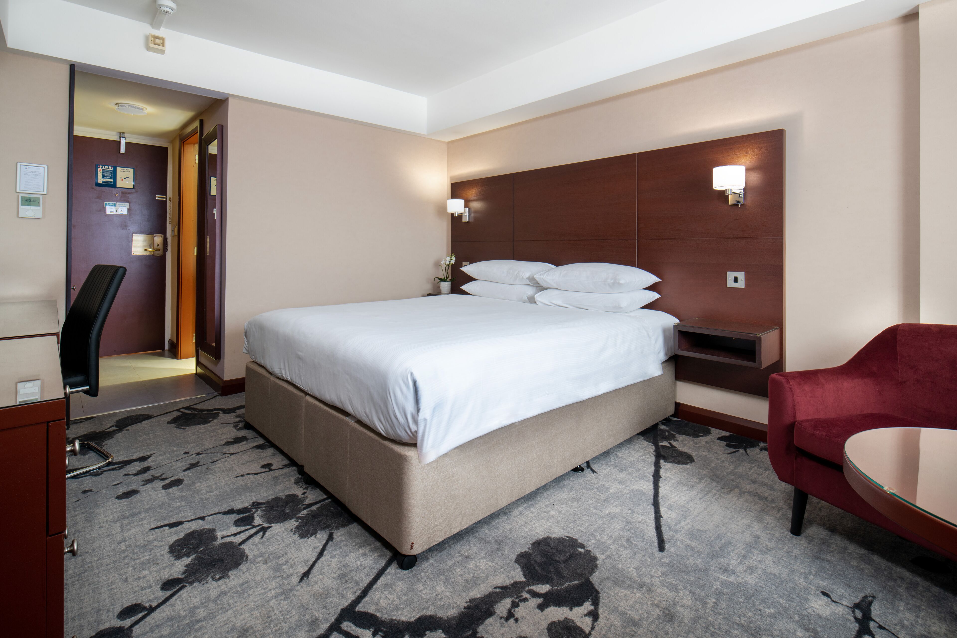 Quarto de hotel com cama, uma cadeira vermelha, mesa de cabeceira e luminária fixada na parede.