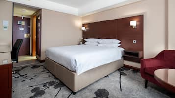 Quarto de hotel com cama, uma cadeira vermelha, mesa de cabeceira e luminária fixada na parede.