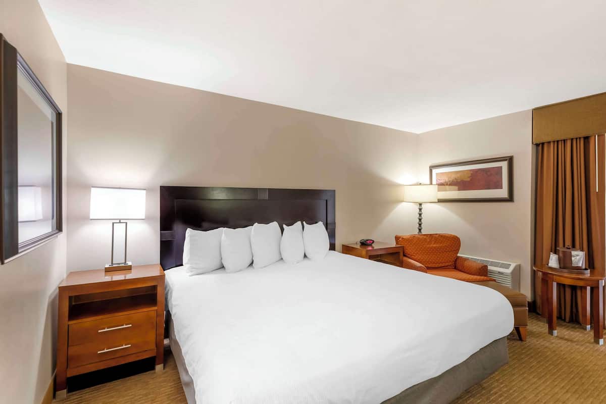 Quarto de hotel com uma cama grande, mesa de cabeceira com gavetas, uma cadeira, abajur e um quadro na parede.