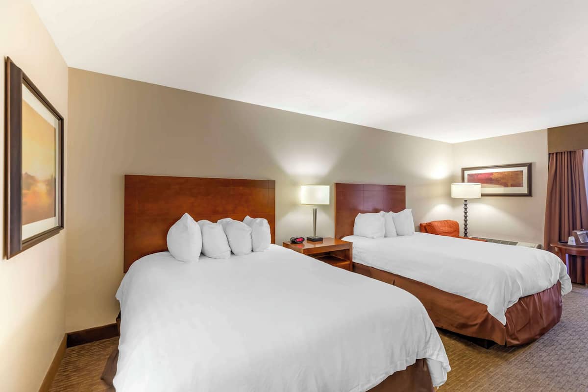 Quarto de hotel com duas camas, cada uma com roupa de cama branca e cabeceiras de madeira. Há mesas de cabeceira com luminárias, um quadro emoldurado na parede e piso coberto de carpete.