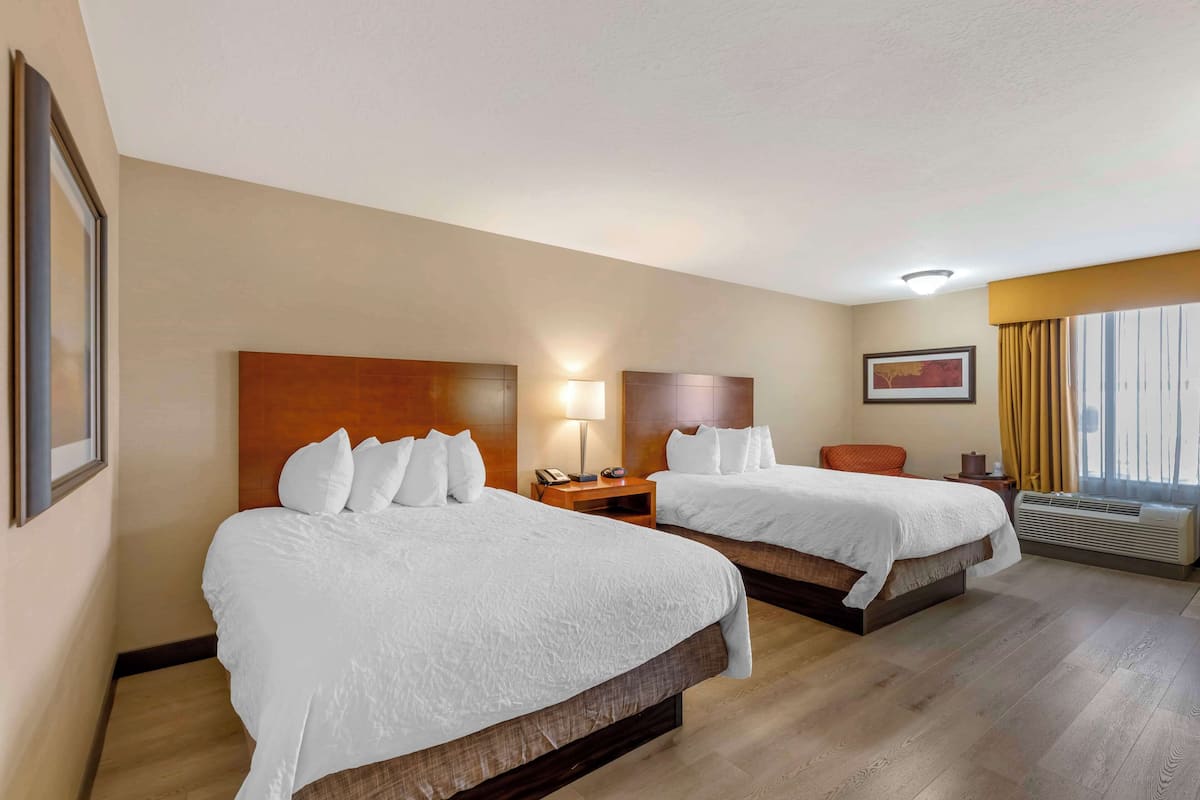 Quarto de hotel com duas camas, uma mesinha de cabeceira com abajur, um quadro na parede e uma janela com cortinas.