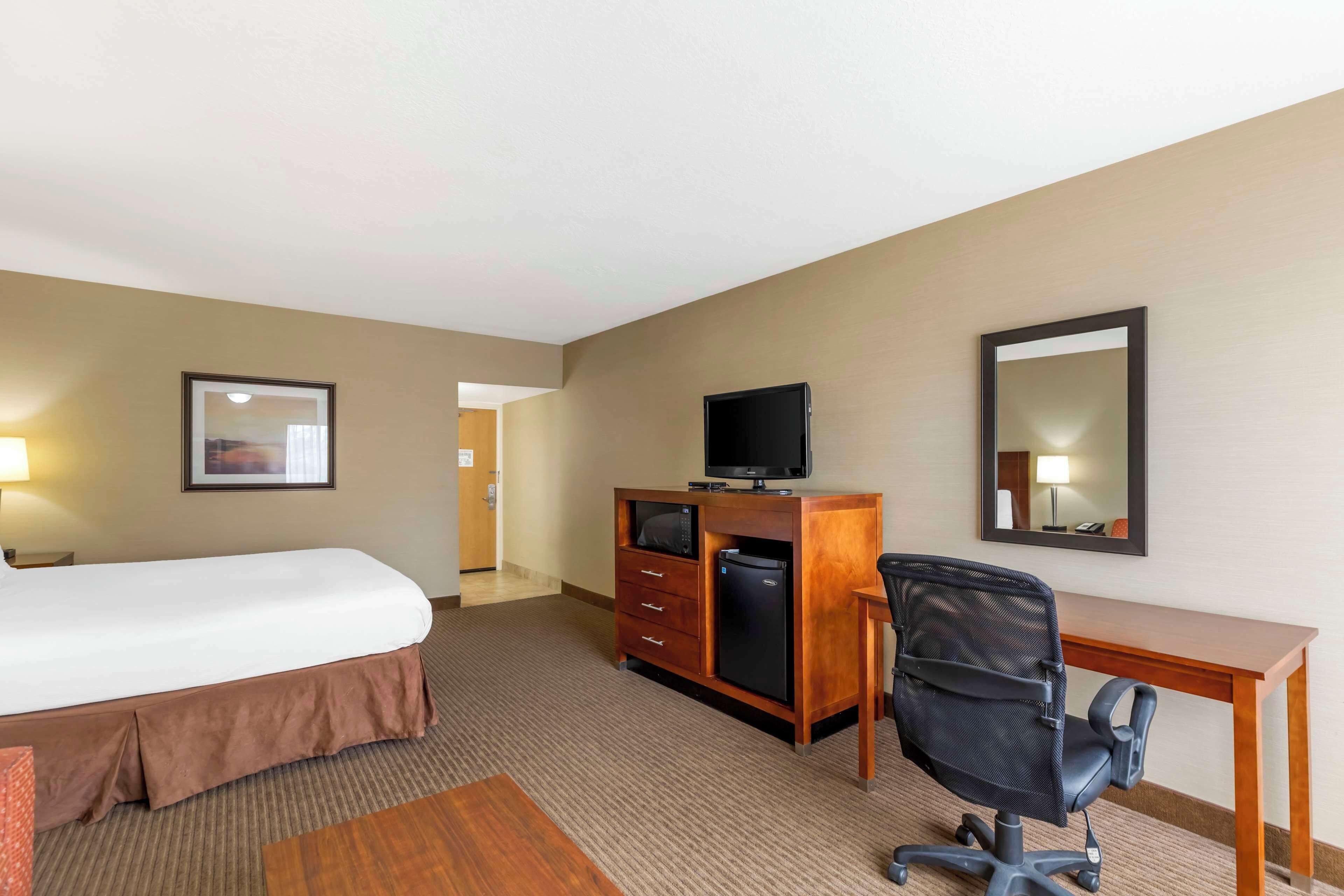 Quarto de hotel com cama, escrivaninha com cadeira, televisão, minibar e espelho.