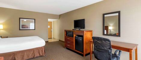 Quarto de hotel com cama, escrivaninha com cadeira, televisão, minibar e espelho.