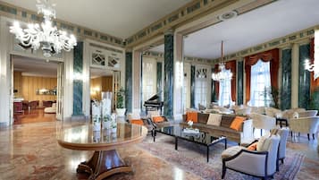 Um hall amplo com um lustre imponente, um piano e uma área de estar com sofás e poltronas.