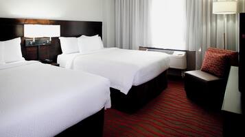 Quarto de hotel com duas camas, um tapete vermelho, uma mesa de cabeceira com abajur e uma cadeira vermelha.