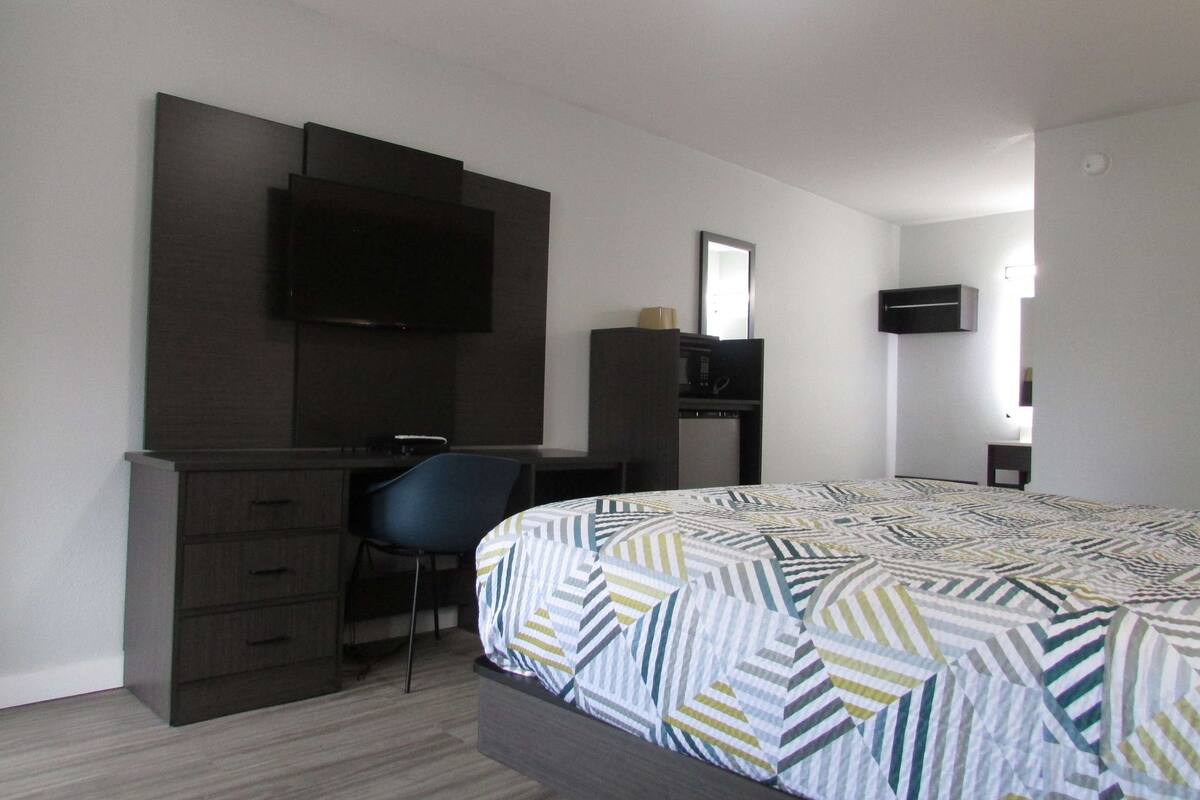 Quarto de hotel com cama, escrivaninha, cadeira, televisão e espelho.