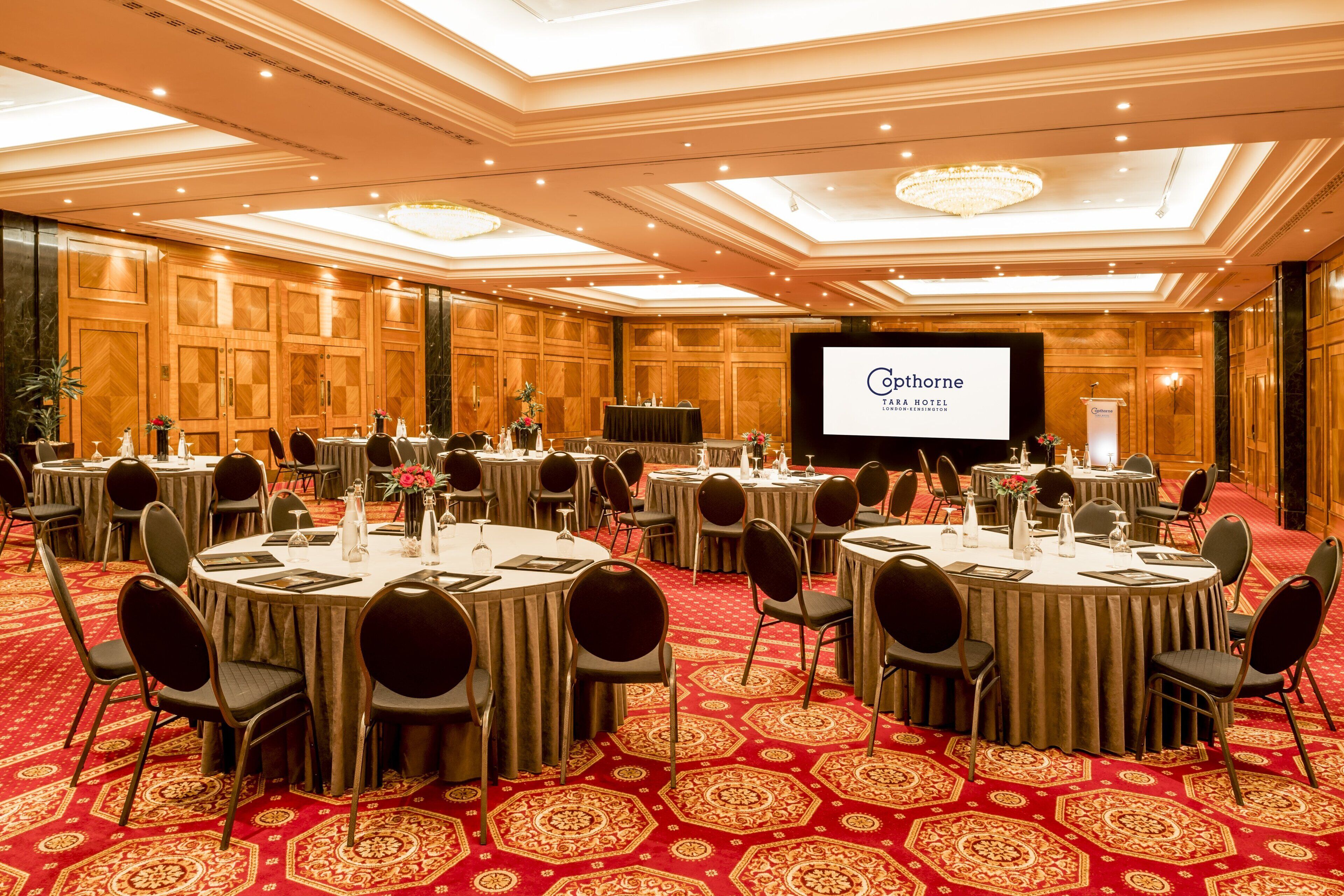 Sala de conferência com várias mesas redondas, cada uma com cadeiras, equipada com porta-nomes e garrafas de água. Uma tela grande exibe o nome do hotel.