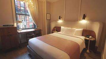 Quarto de hotel com uma cama grande, uma escrivaninha, uma cadeira, uma mesinha e uma janela com cortinas.