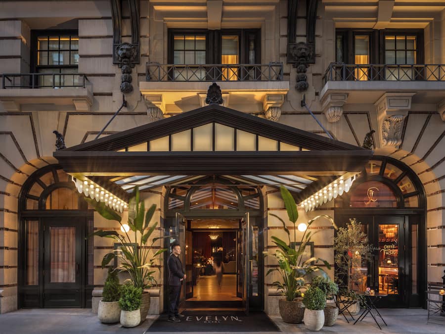 Entrada de hotel com acesso coberto, plantas em vasos e placa de restaurante visível.