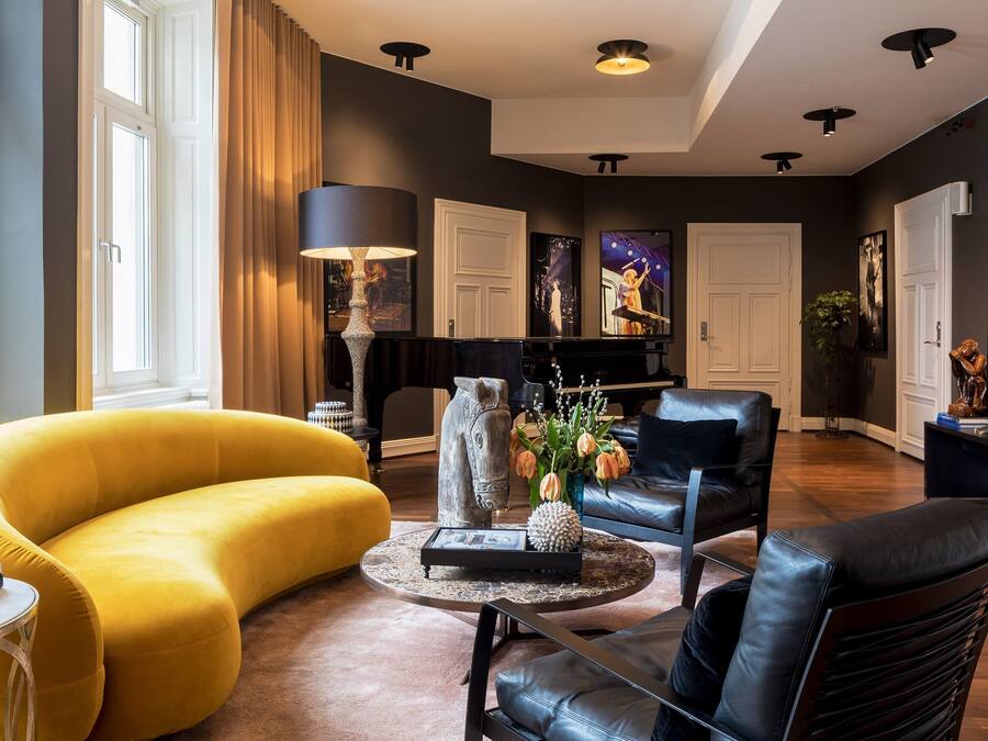 Uma sala de estar com um sofá amarelo, uma poltrona de couro preto, um piano de cauda e uma grande janela com cortinas.