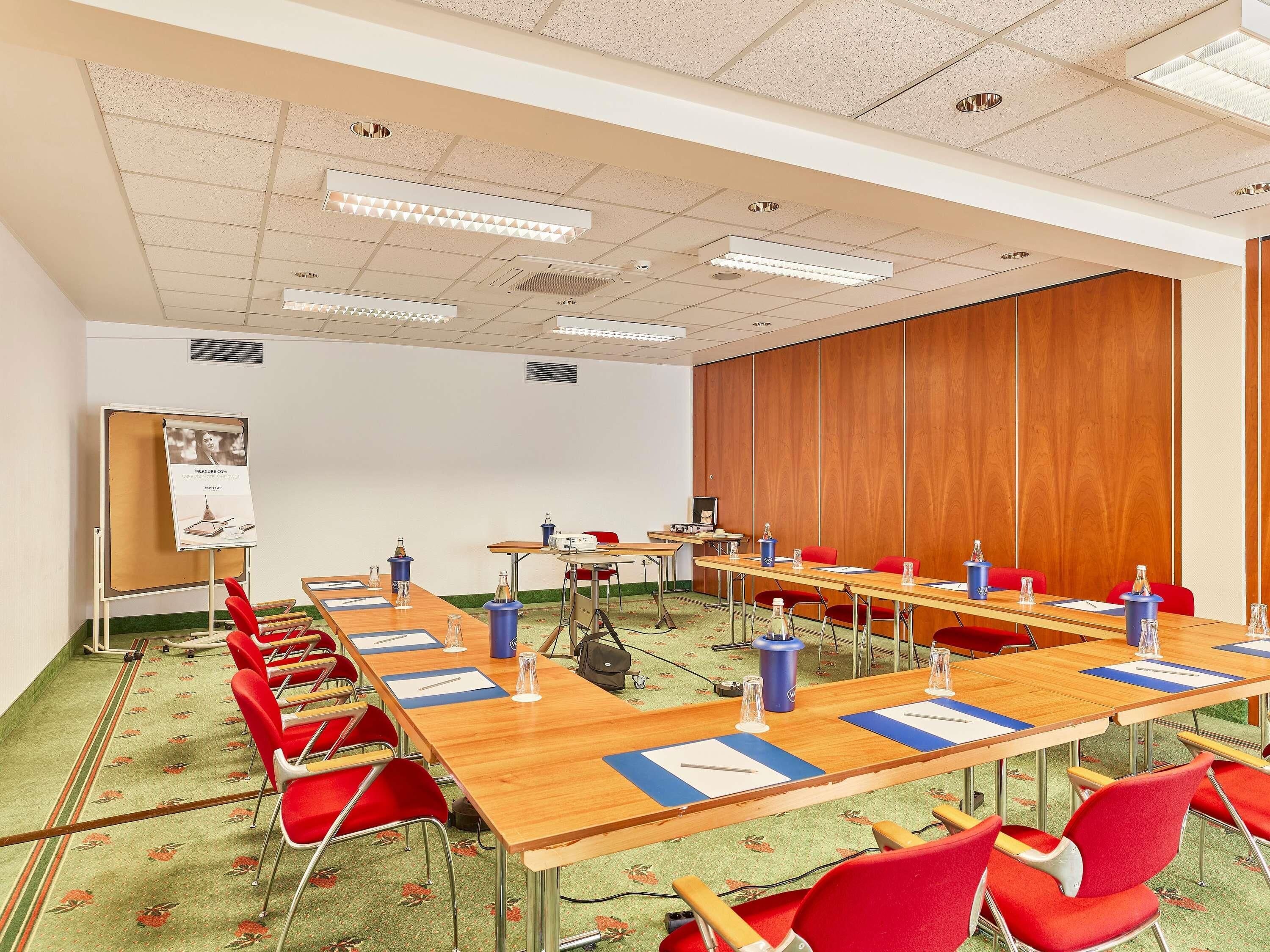 Uma sala de conferência com uma mesa comprida, cadeiras vermelhas, um quadro branco e um painel divisório de madeira.