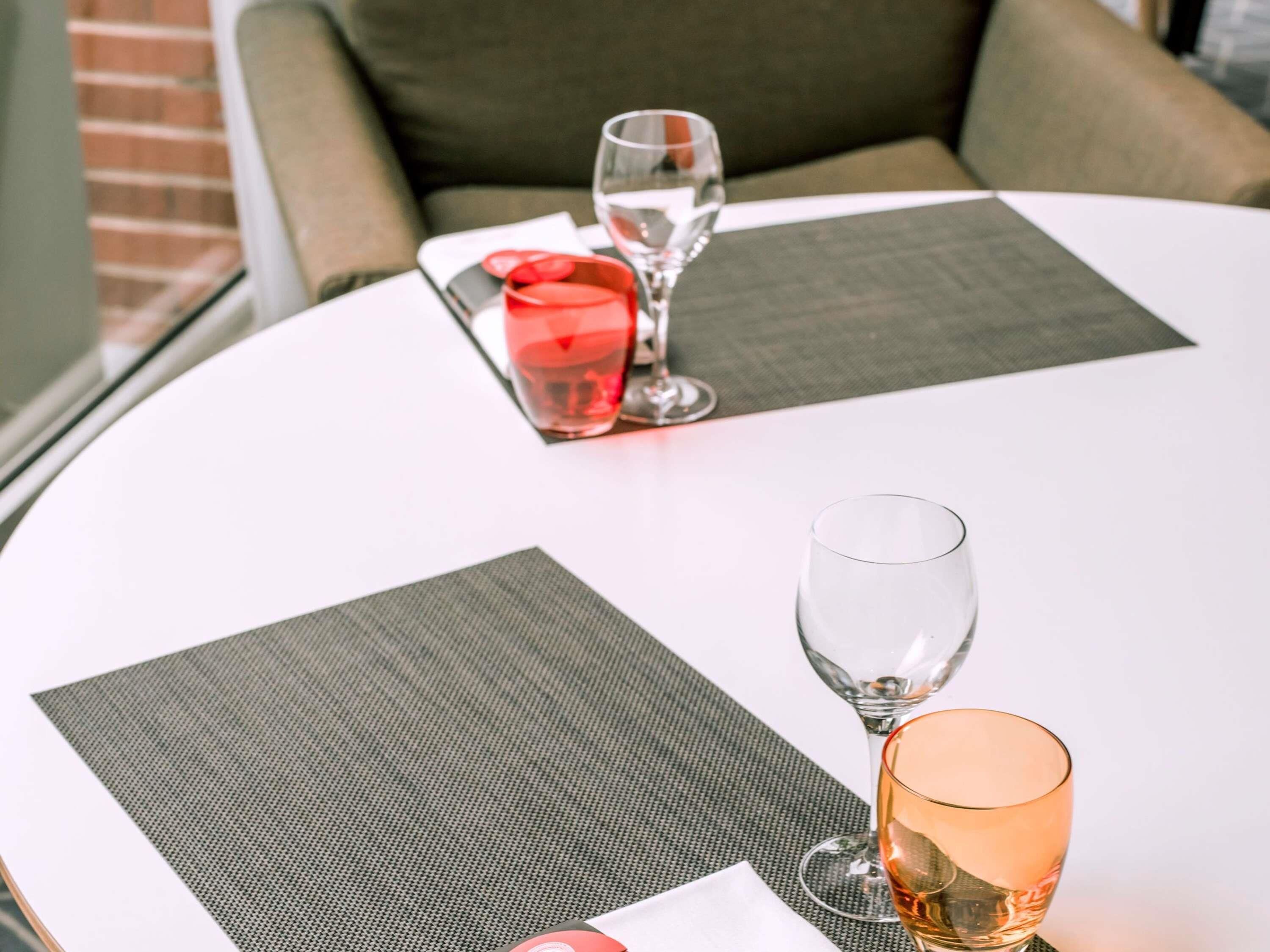 Uma mesa redonda com duas taças de vinho e uma caneca vermelha, disposta com um guardanapo branco e um apoio para prato.