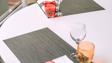 Uma mesa redonda com duas taças de vinho e uma caneca vermelha, disposta com um guardanapo branco e um apoio para prato.