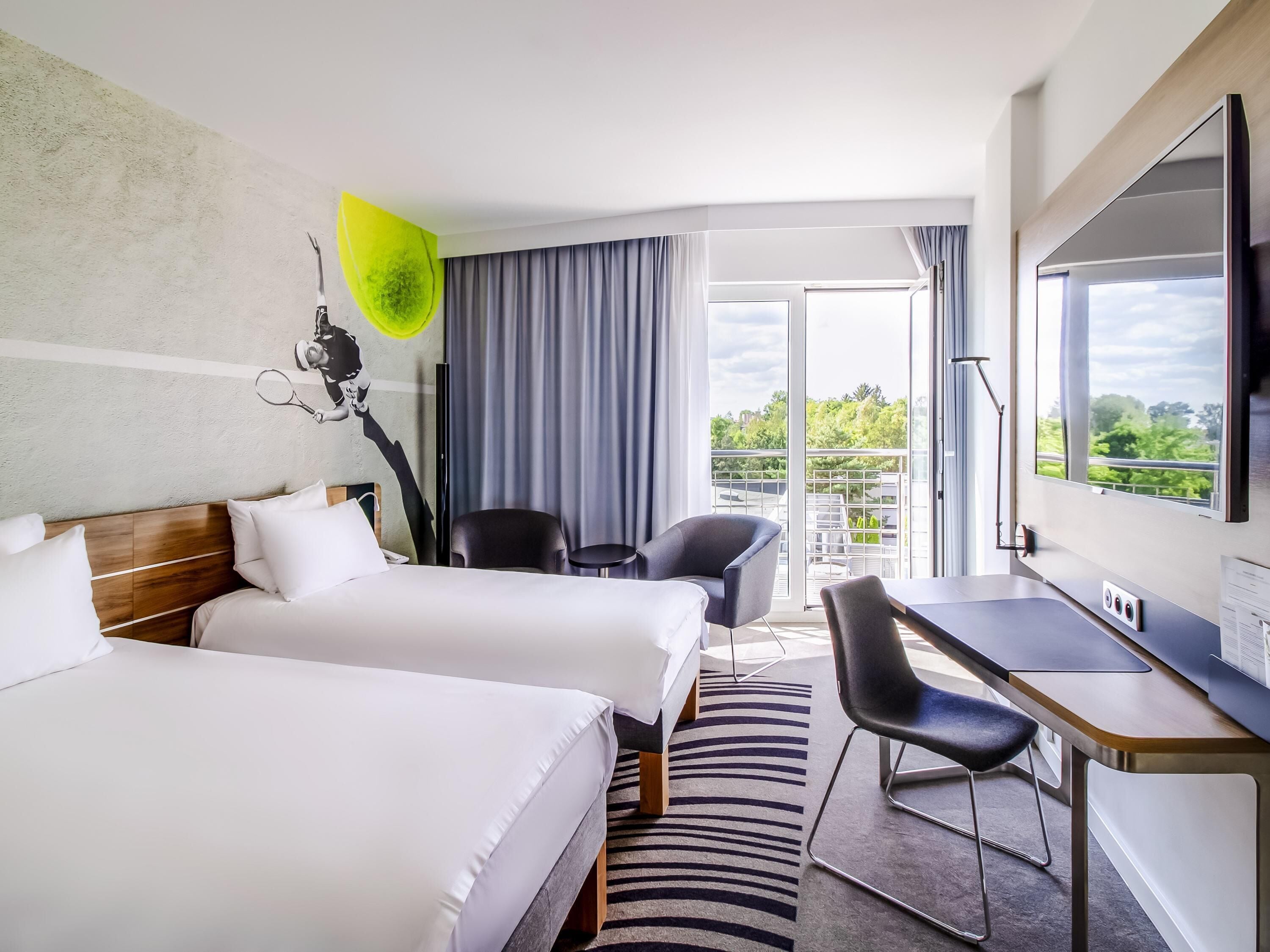 Quarto de hotel com duas camas, uma escrivaninha, uma cadeira, uma televisão e varanda com vista.