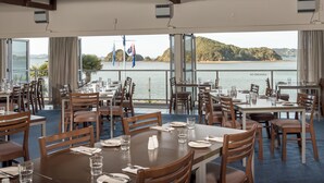 Restaurante com mesas preparadas para refeições, grandes janelas com vista para o mar e colinas, e bandeiras penduradas na parte externa.