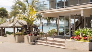 Um hotel moderno com fachada de vidro e vista para a água, área externa coberta com plantas e uma escadaria que leva à entrada.