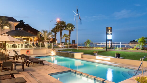 Piscina com espreguiçadeiras e guarda-sóis, um mastro e uma placa de sinalização ao fundo.