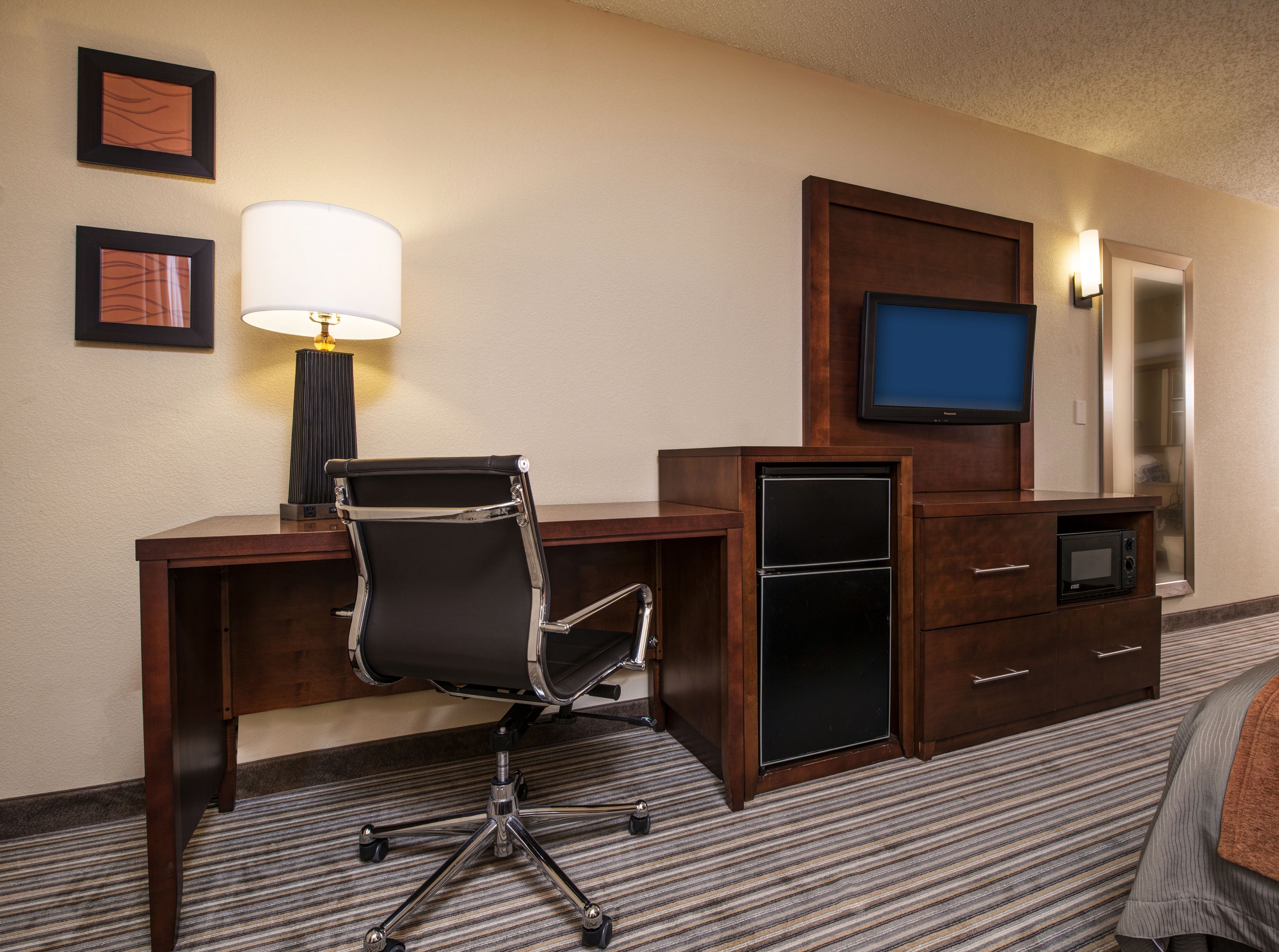 Quarto de hotel com escrivaninha de madeira, cadeira de escritório preta, luminária, televisão, micro-ondas e cômoda.