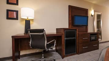 Quarto de hotel com escrivaninha de madeira, cadeira de escritório preta, luminária, televisão, micro-ondas e cômoda.
