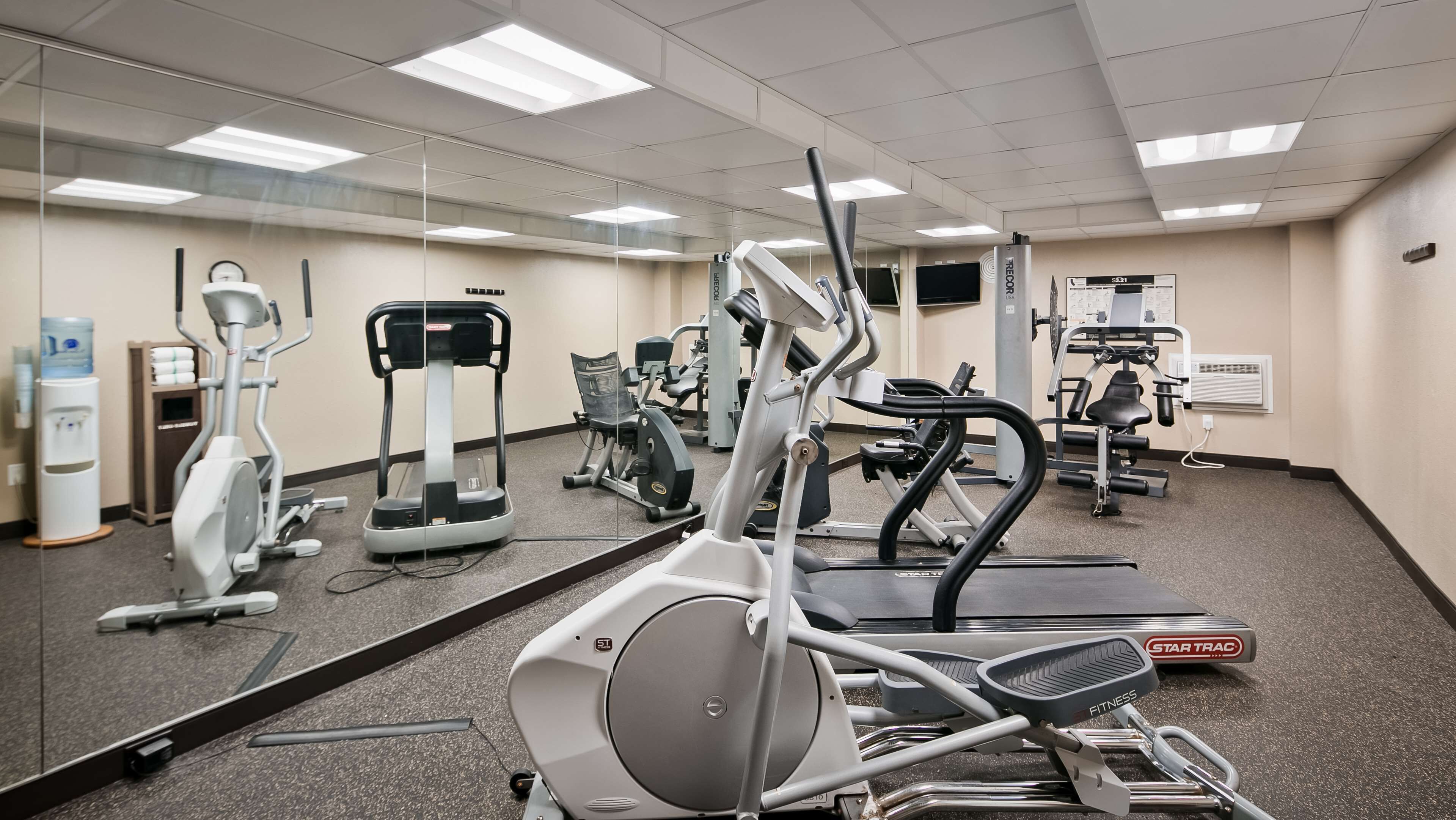 Uma academia com diversos aparelhos de ginástica, um bebedouro e um espelho refletindo os equipamentos.
