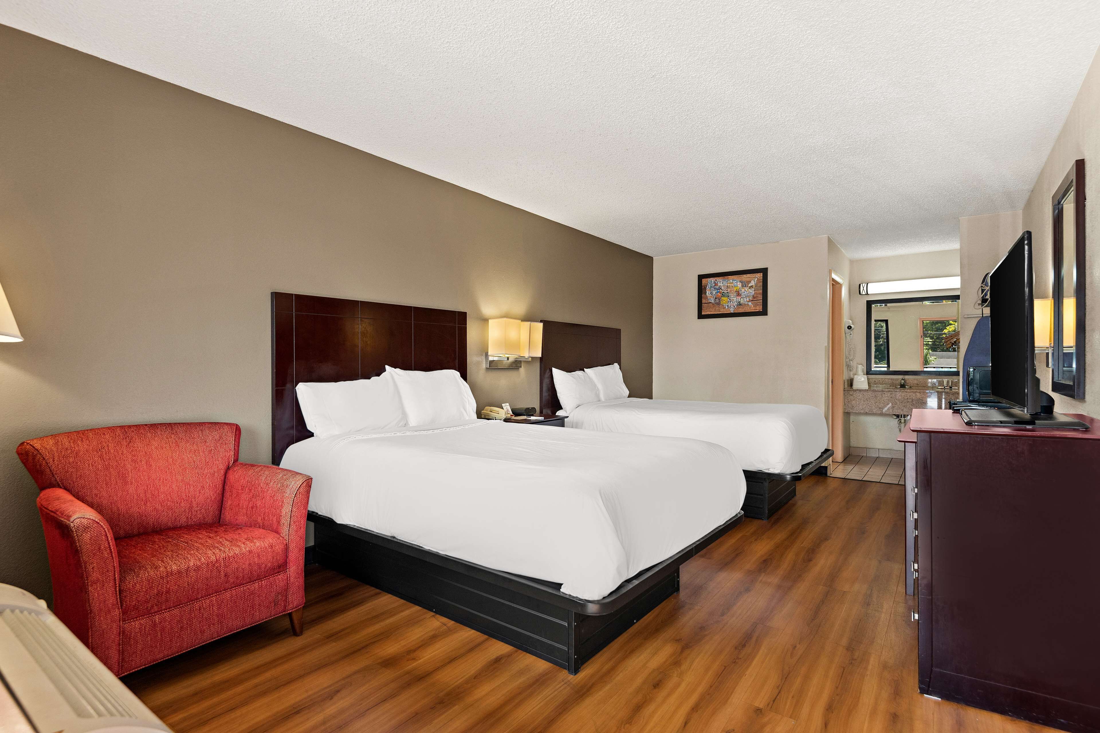 Quarto de hotel com duas camas, uma poltrona vermelha, uma escrivaninha com televisão e um banheiro visível por uma porta aberta.