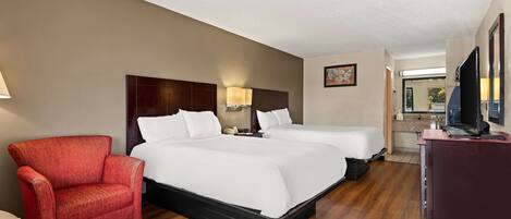 Quarto de hotel com duas camas, uma poltrona vermelha, uma escrivaninha com televisão e um banheiro visível por uma porta aberta.