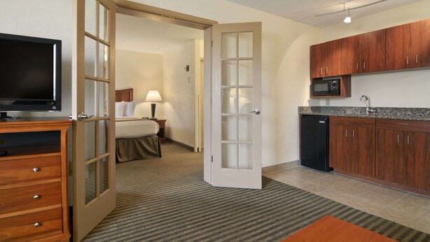 Quarto de hotel com cama, mesa de cabeceira, luminária, televisão, cómoda de madeira e cozinha equipada com micro-ondas e máquina de lavar louça.