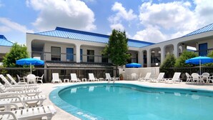 Uma área de piscina com cadeiras de lounge brancas e guarda-sóis azuis, um edifício com telhado azul e céu claro.