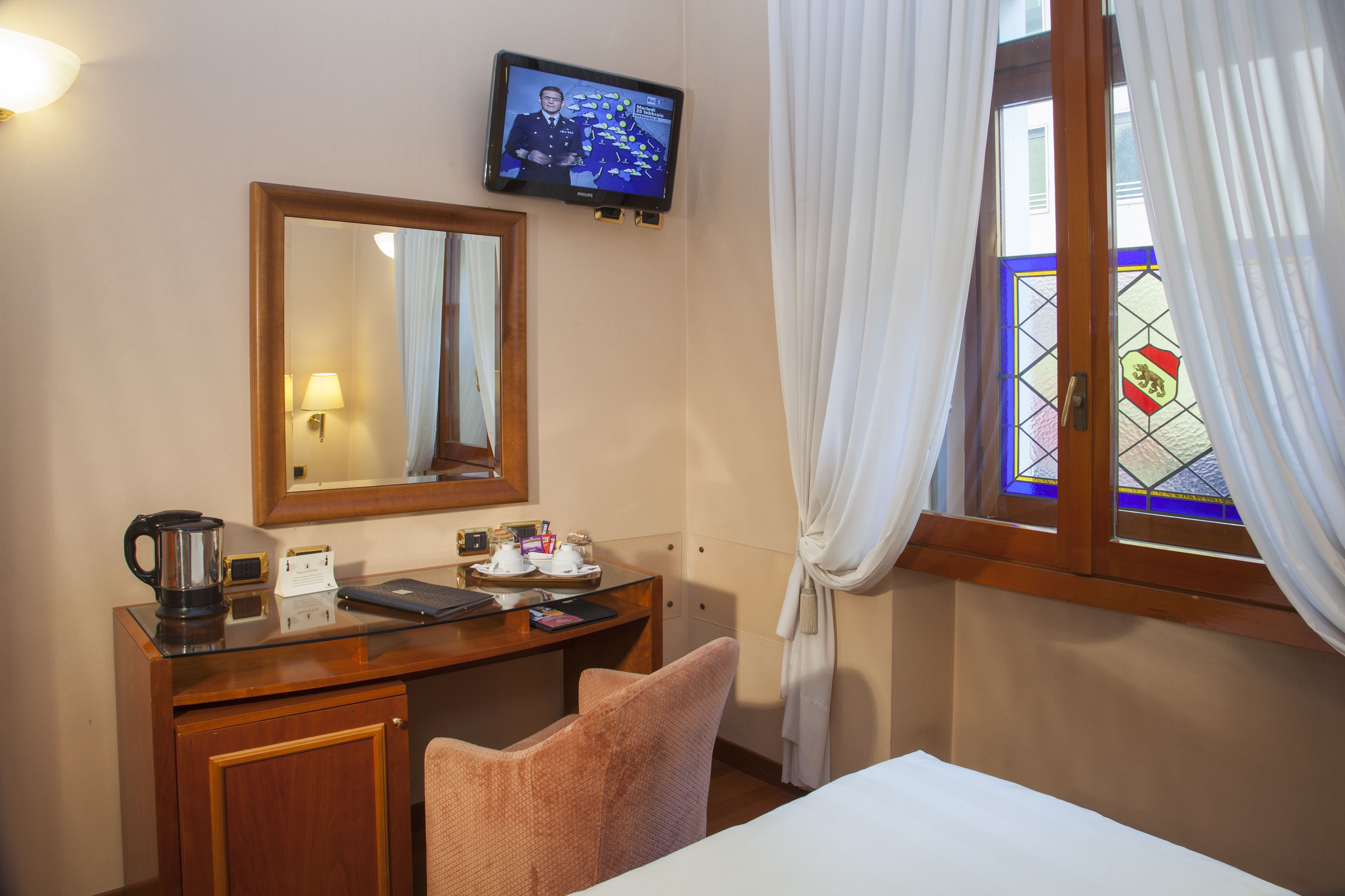 Quarto de hotel com uma escrivaninha de madeira, televisão fixada acima, cama com roupa de cama branca, janela com cortinas e um espelho na parede.
