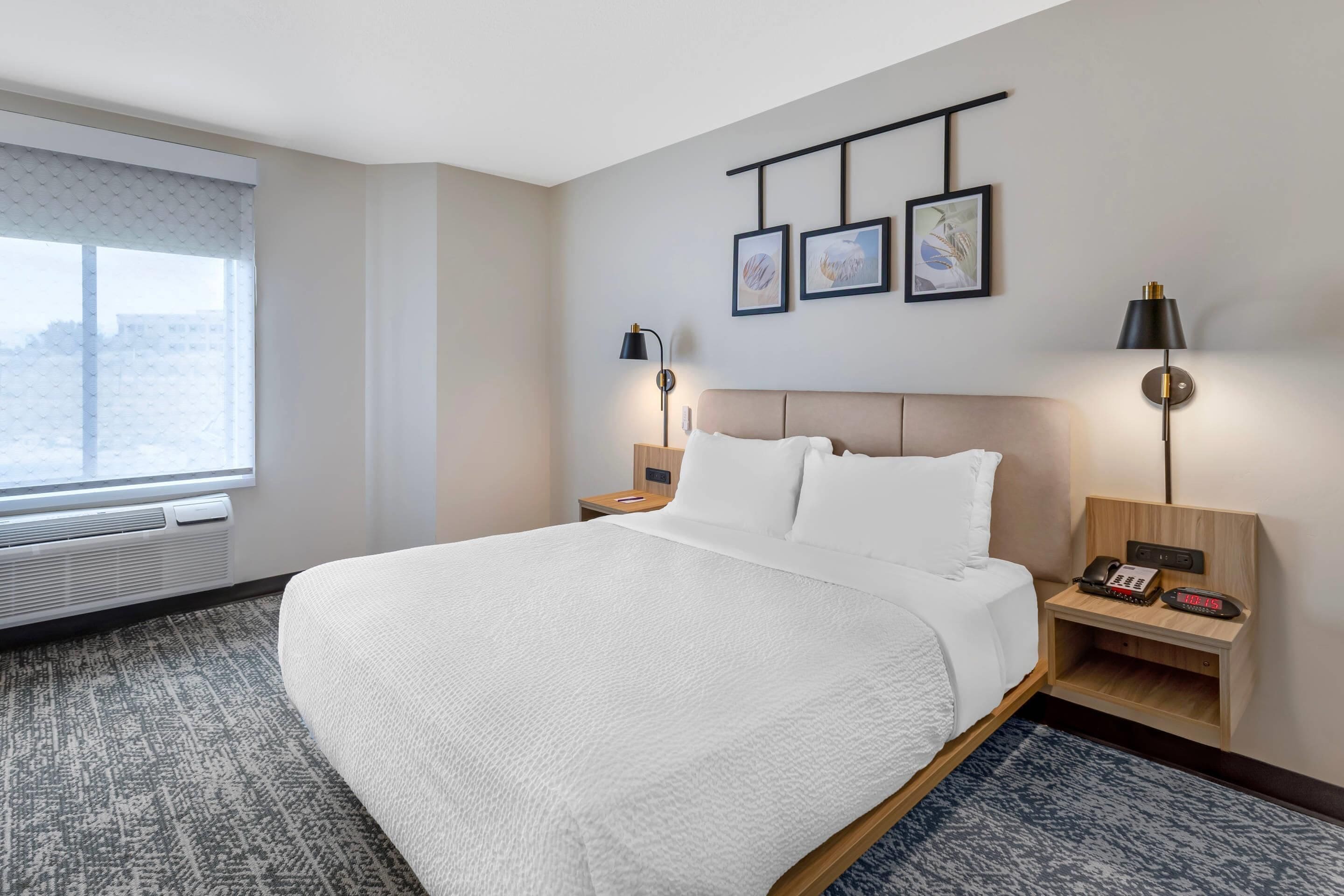 Quarto de hotel com uma cama grande, mesas de cabeceira com abajures, uma janela com persianas e obras de arte na parede.