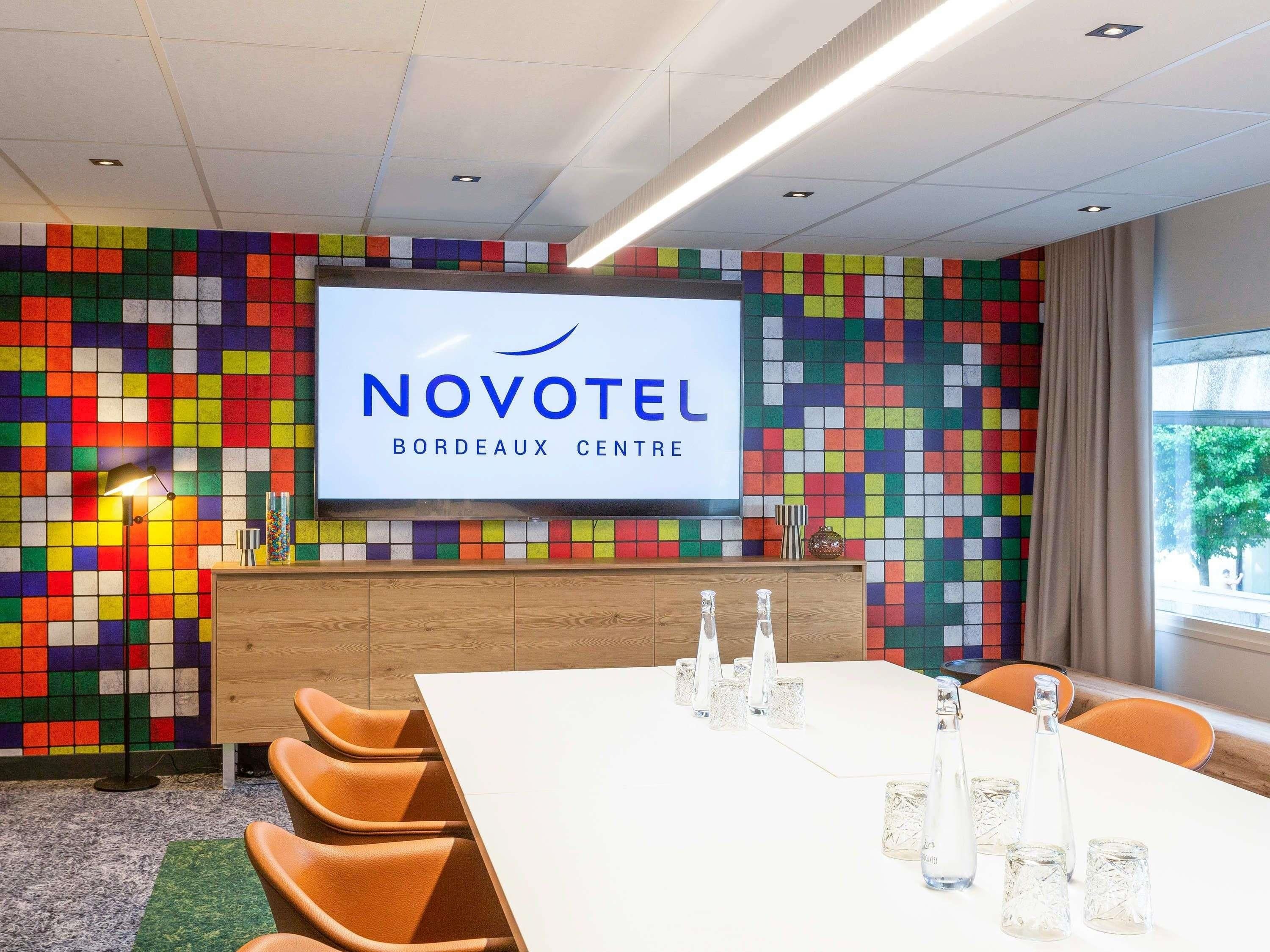 Quarto de hotel com parede em mosaico colorido, uma grande tela de TV exibindo o logotipo do Novotel Bordeaux Centre, um armário de madeira, uma mesa branca com cadeiras e uma janela com vista.