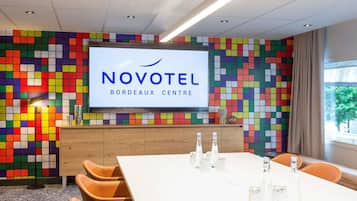 Quarto de hotel com parede em mosaico colorido, uma grande tela de TV exibindo o logotipo do Novotel Bordeaux Centre, um armário de madeira, uma mesa branca com cadeiras e uma janela com vista.