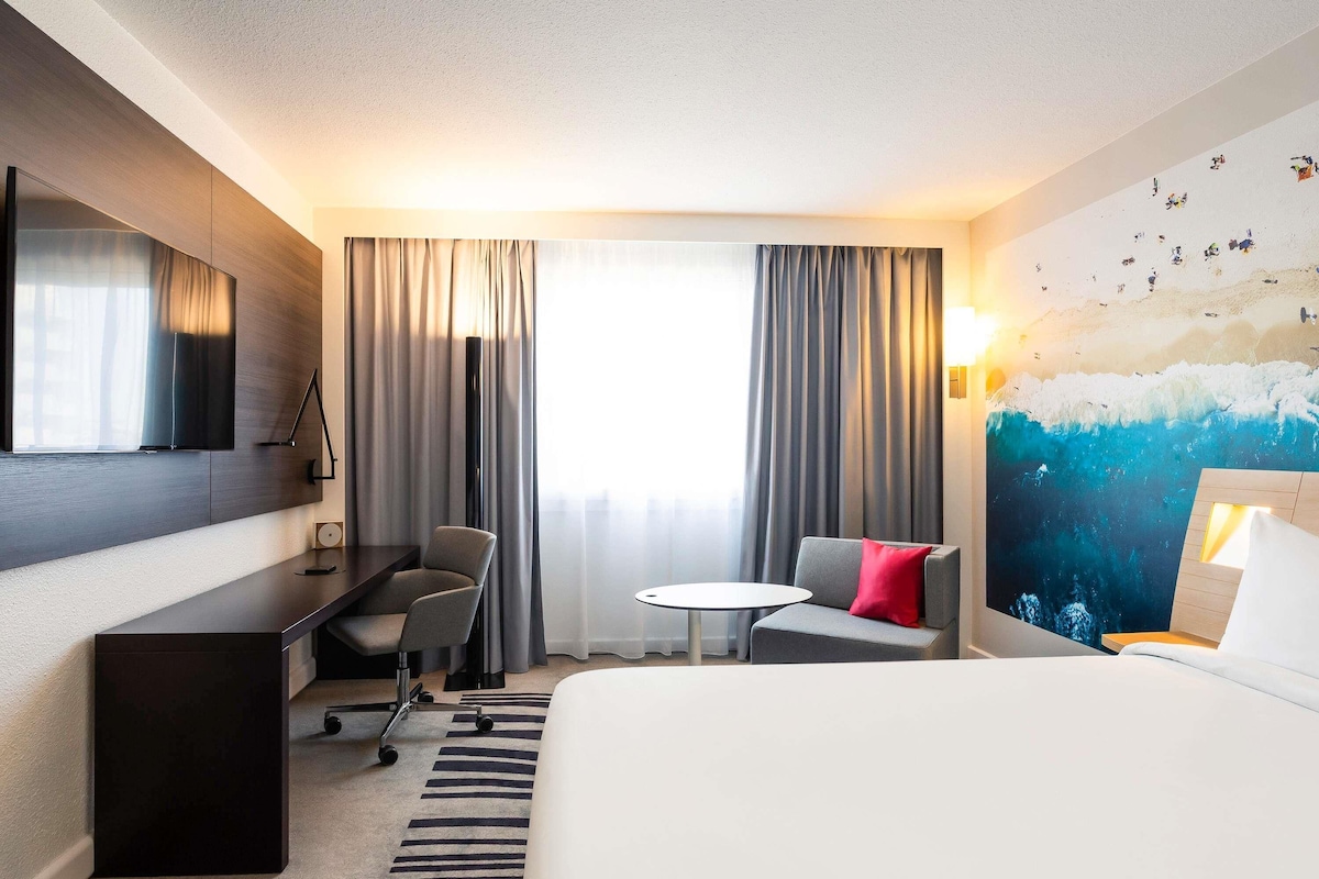 Quarto de hotel com cama, escrivaninha, cadeira, mesa pequena e janela grande com cortinas.