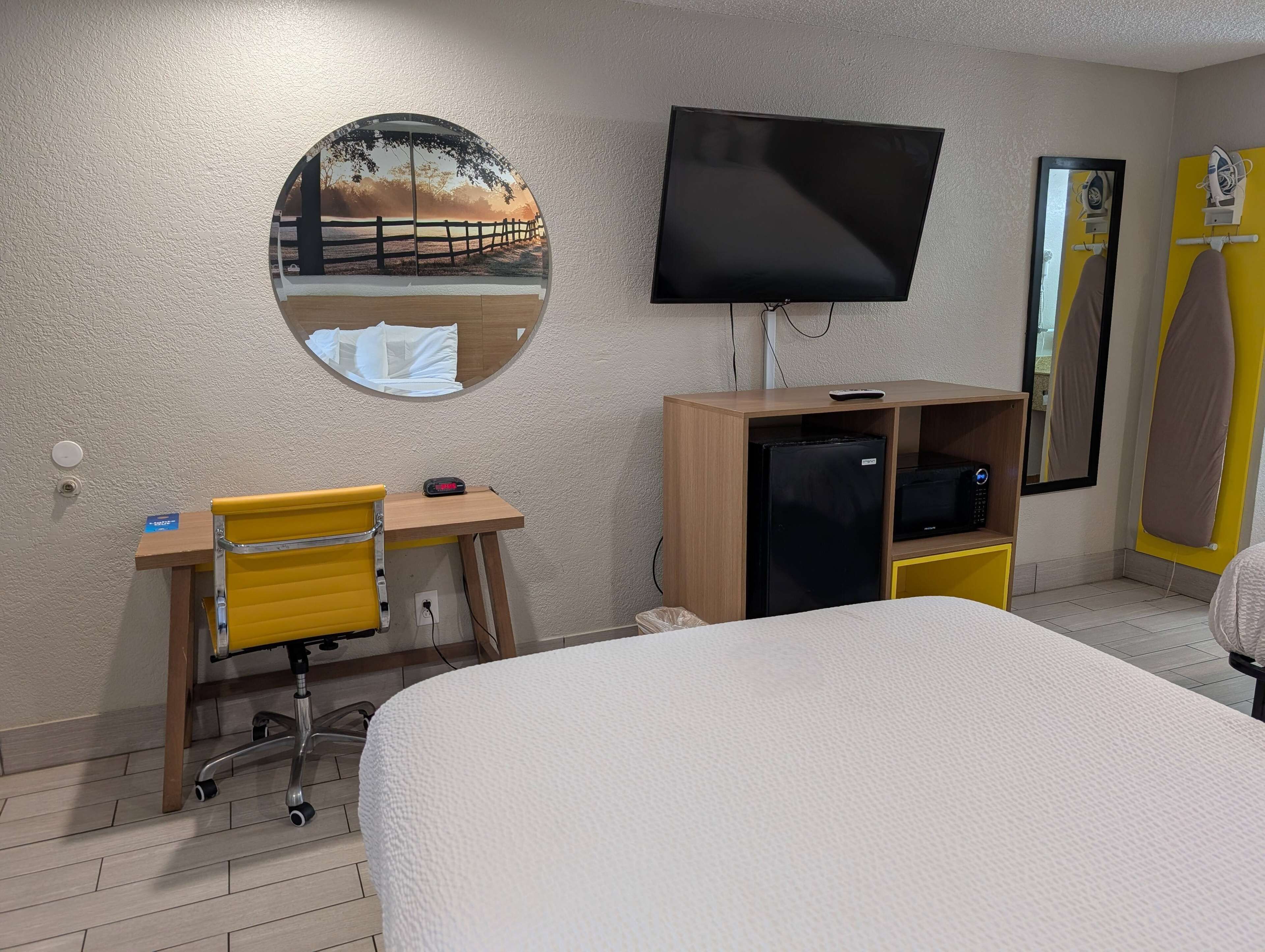 Quarto de hotel com cama, escrivaninha com cadeira, televisão, espelho e pequeno frigorífico.