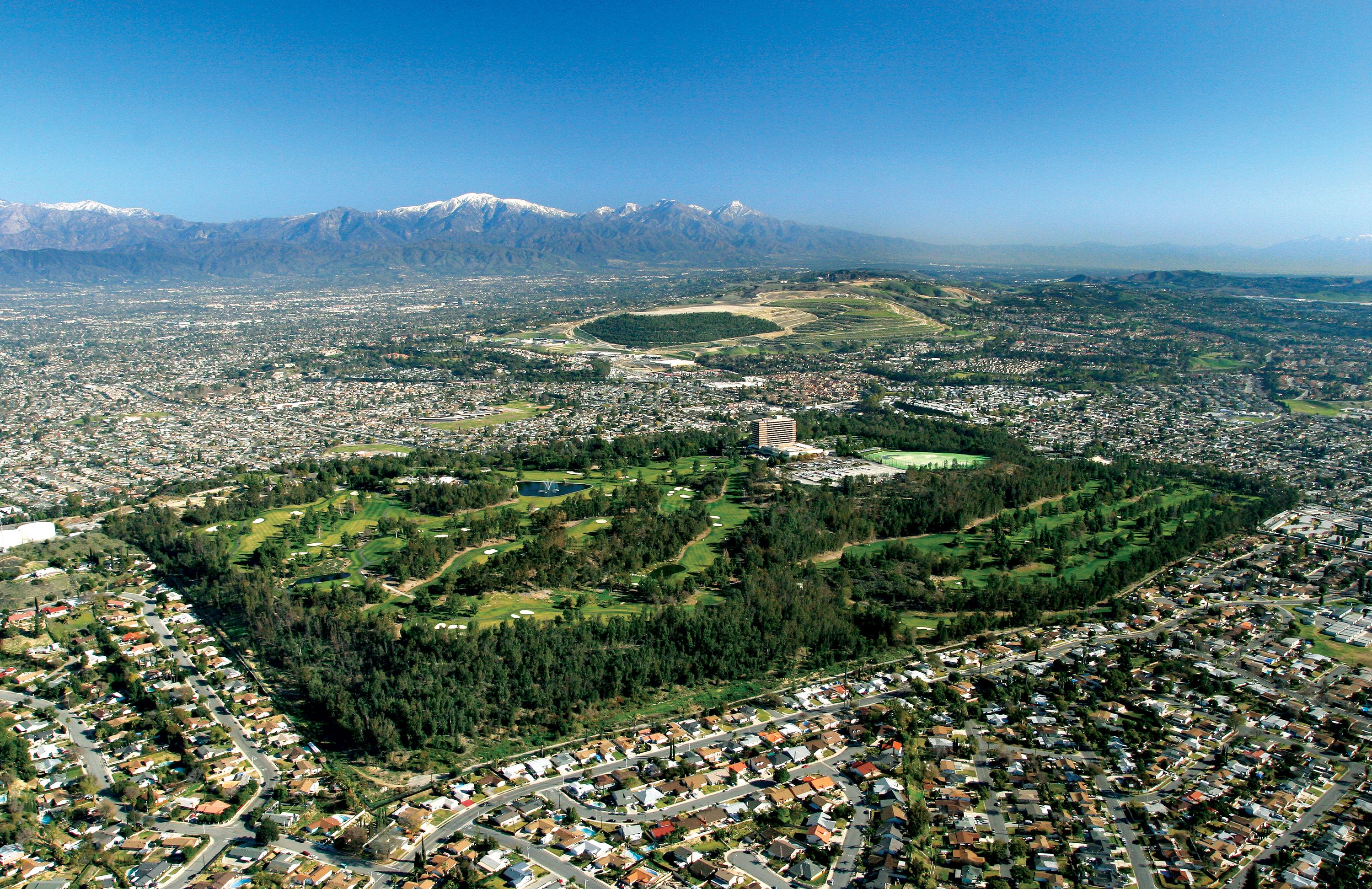 Vista aérea de uma cidade com um grande parque no centro, rodeada por áreas residenciais e montanhas ao fundo.